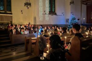 A candlelit carol service in the Lady Margaret Hall chapel. The choir is gathered in the front pews, holding lit candles and singing, while a speaker stands at the lectern. Congregants fill the pews, also holding candles. A lit Christmas tree glows to the right of the altar. The atmosphere is warm and reflective, with stained glass window reflections cast on the wall.